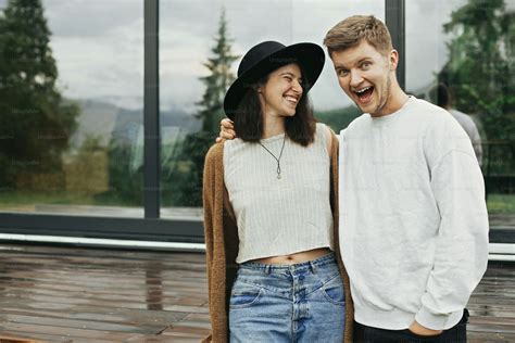 세련된 힙스터 커플은 나무 베란다에서 웃으며 산의 현대적인 오두막에서 휴식을 취하고 있습니다 테라스와 큰 창문을 배경으로 웃고