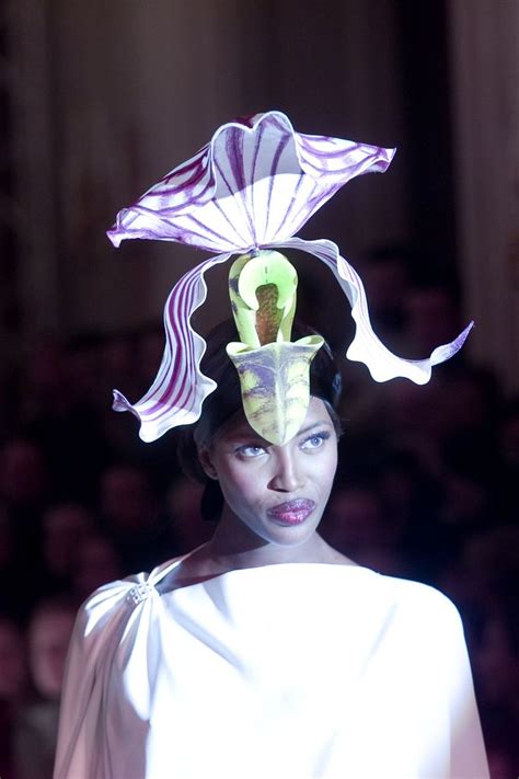 A Woman In A White Dress And Hat With Flowers On Her Head At A Fashion Show