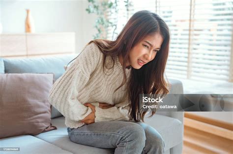복통을 앓고 있는 젊은 아시아 여성은 집 거실의 소파에 앉아 복통 위염 고통스러운 생리 개념을 앓고 있다 위통에 대한 스톡 사진