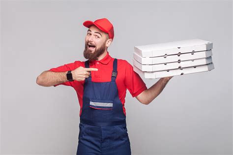 파란색 유니폼을 입고 수염과 젊은 행복 배달 남자와 빨간 T셔츠 서 들고 가리키는 회색 배경에 골 판지 피자 상자 더미를 손가락 유머에 대한 스톡 사진 및 기타 이미지