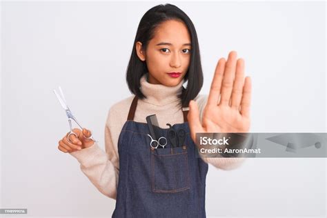중국 미용사 여자 에이프런 을 입고 가위를 들고 고립 된 흰색 배경 위에 열린 손으로 정지 표지판을하고 진지하고 자신감있는 표정