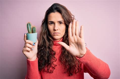 진지하고 자신감있는 표정으로 정지 표지판을하고 열린 손으로 고립 된 분홍색 배경 위에 작은 선인장을 들고 곱슬 머리를 가진 젊은