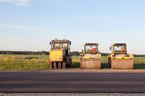 도로 수리 중 트랙 측면에 서있는 세 개의 큰 노란색 아스팔트 포장 롤러와 여름에 푸른 하늘과 구름이있는 풍경에 검은 색 새로운 아스팔트를 누워 거리에 대한 스톡 사진 및