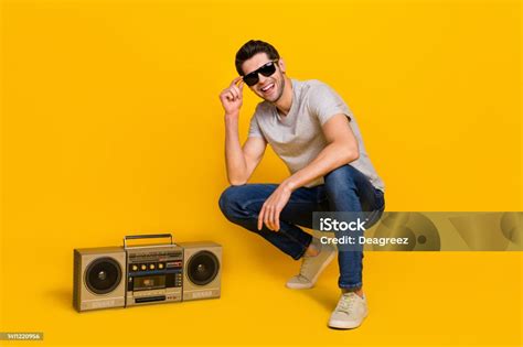 붐박스 근처에 앉아있는 멋진 브루넷 젊은 남자의 풀 사이즈 사진 노란색 배경에 고립 된 티셔츠 안경 청바지 운동화를 착용하십시오