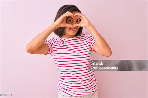 줄무늬 티셔츠를 입은 젊은 아름다운 여성은 사인을 통해 보고 미소 와 손과 손가락으로 심장 모양을하고 고립 된 분홍색 배경 위에