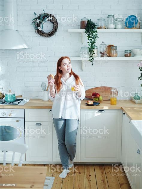 아름 다운 정통 젊은 여 자가 먹는 빵을 즐기는 빨간 머리와 아침 식사와 낮에는 오렌지 주스를 마시는 부엌에서 카운터에 기대어 햇빛에 꿈 몸 긍정주의에 대한 스톡 사진 및