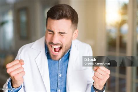 클리닉에서 젊은 잘생긴 의사 남자 매우 행복 하 고 승리 웃 고 성공에 대 한 비명 팔과 승자 제스처를 하 고 흥분 축 하 개념입니다 건강관리와 의술에 대한 스톡 사진 및