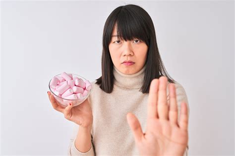 아름답고 고립된 흰색 배경 위에 마시멜로가 있는 그릇을 들고 있는 아름다운 중국 여성은 진지하고 자신감있는 표정 방어 제스처로 정지 표지판을 하는 오픈 핸드로 고립 된 흰색