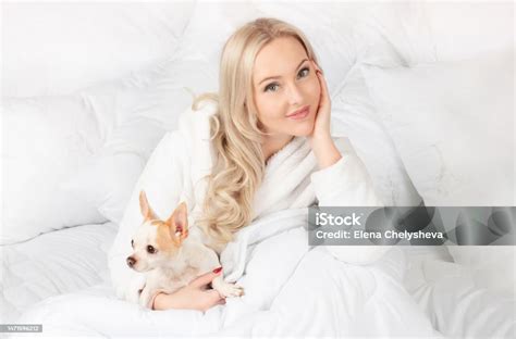 테리 흰 가운을 입고 개와 함께 금발 소녀가 담요와 베개가있는 침대에 앉아 있습니다 35 39세에 대한 스톡 사진 및 기타