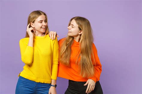 두 웃는 젊은 금발 쌍둥이 자매는 밝은 화려한 옷 서 서 파스텔 보라색 파란색 배경에 고립 된 서로를 보고 사람들 가족 라이프