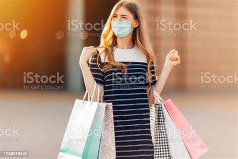 매력적인 세련된 젊은 여성은 쇼핑 가방을 들고 쇼핑 센터를 떠나 그녀의 얼굴에 의료 보호 마스크를 입고 검역 코로나바이러스