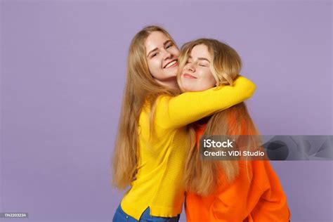 파스텔 바이올렛 블루 벽 배경에 고립 된 찾고 카메라를 포옹 하는 생생한 다채로운 의류에서 두 귀여운 웃는 젊은 금발 쌍둥이 자매 여자 사람들 가족 라이프 스타일 개념입니다