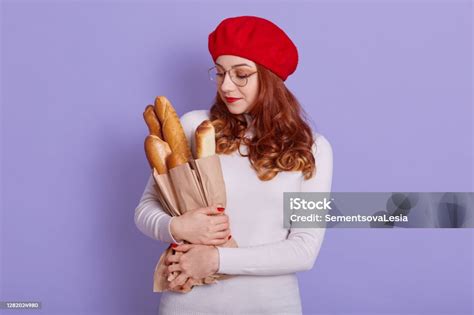 라일락 배경에 빵이 달린 종이 봉투를 들고 있는 아름다운 젊은 여성의 초상화 흰색 캐주얼 셔츠와 빨간 베레모를 입은 신선한 향기로운 롱 로프를 가진 소녀의 초상화는 물결