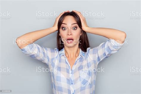 안 돼요 활짝 열린된 입과 회색 배경에 고립 된 두 손으로 머리를만 지는 체크 무늬 셔츠에 눈을 가진 예쁜 놀 충격 여자의