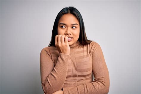 젊은 아름 다운 아시아 여자 캐주얼 터틀넥 스웨터 를 입고 흰색 배경 위에 서 스트레스와 긴장 한 입 물고 손톱에 손으로 불안