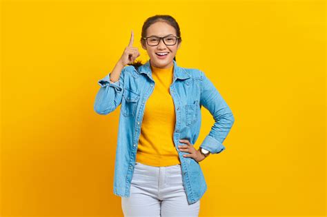 데님 옷을 입고 웃는 젊은 아시아 여성 학생의 초상화는 복사 공간에서 손가락을 가리키며 노란색 배경에 고립 된 광고 제품을 보여