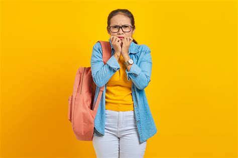 배낭이있는 데님 복장을 입은 아름다운 젊은 아시아 학생은 노란색 배경에 손톱을 물어 뜯는 입에 손으로 스트레스를 받고 긴장한