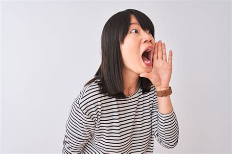 고립 된 흰색 배경 위에 검은 줄무늬 티셔츠를 입은 젊은 아름다운 중국 여성이 입에 손을 대고 큰 소리로 비명을 지르고 있습니다
