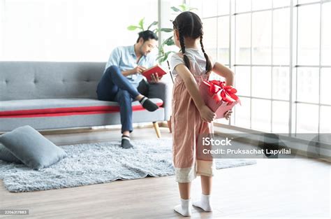 아시아 귀여운 어린 딸이 등 뒤로 포장 된 선물 상자를 들고 숨기고 아빠를 놀라게하고 아버지의 날이나 생일을 맞이하고 소파에 앉아서 책을 읽는 아빠를 맞이합니다 2명에 대한