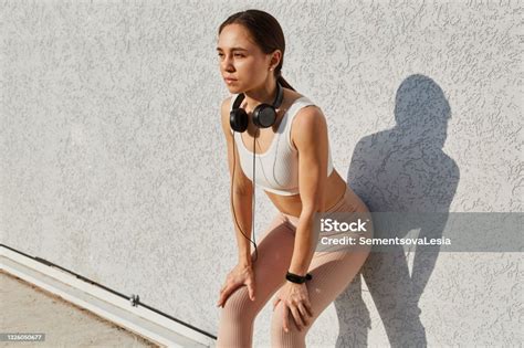 흰색 상의와 베이지 색 레긴을 입고 완벽한 몸매를 가진 스포티 한 여성은 야외에서 회색 벽에 기대어 포즈를 취하고 달리고 헤드폰으로 음악을 듣는 포즈를 취했습니다 25 29