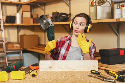 격자 무늬 셔츠에 젊은 갈색 머리 여자 회색 T셔츠 소음 절연 헤드폰 노란색 장갑 다른 도구 나무 테이블 장소에 목공 워크숍에서 힘을 가진 나무를 톱 질 보았다 Diy에