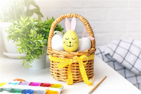 귀여운 토끼 얼굴과 귀가 있는 노란색 계란은 페인트와 브러시가 있는 흰색 테이블에 리본이 있는 바구니에 있습니다 행복한 부활절