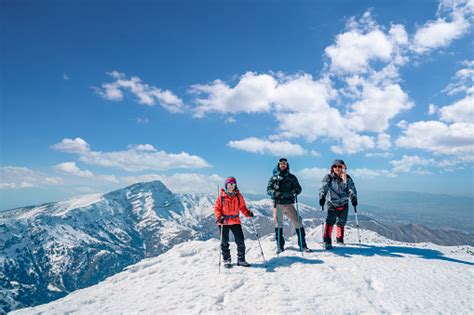 성공적인 산악 등반팀은 겨울에 고지대 눈 덮인 산 정상의 봉우리에 하이킹 기둥을 들고 카메라를 바라보고 있습니다 25 29세에