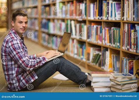 Усмехаясь молодой студент сидя на поле библиотеки используя компьтер книжку Стоковое Изображение