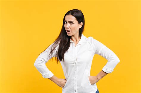 허리에 팔이 Akimbo에 서 흰색 캐주얼 셔츠에 화가 엄격한 젊은 여자의 초상화 맹세 스튜디오 노란색 오렌지 배경에 고립 사람들 라이프 스타일 개념입니다 복사 공간을 모의