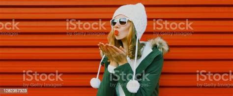 빨간 배경에 폼 폼과 흰색 모자를 입고 달콤한 공기 키스를 보내는 빨간 입술을 불고 젊은 여자의 겨울 초상화 20 29세에 대한 스톡 사진 및 기타 이미지 Istock