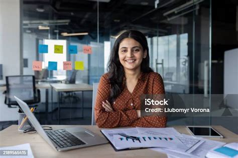 차트와 노트북이 있는 현대적인 사무실의 책상에 앉아 팔짱을 낀 자신감 넘치는 라틴 아메리카 사업가가 성공과 리더십을 반영합니다