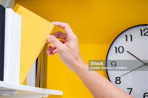 여가 시간에 독서를 통한 자기 계발 남자 손은 노란 벽에 시계가 있는 거실의 책장에서 노란 책을 고르고 따고 있다 Book