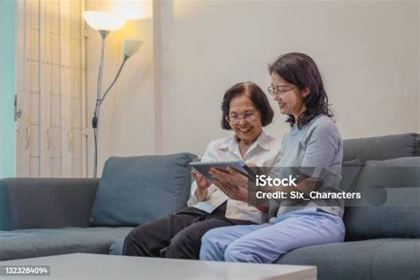 아시아 의 딸과 선임 어머니는 집에서 거실에서 소파에 앉아있는 동안 디지털 태블릿 컴퓨터와 함께 일 노인에 대한 스톡 사진 및
