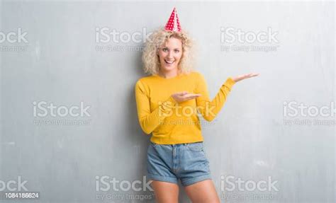 그런 지 회색 벽 입고 생일에 젊은 금발의 여 자가 깜짝 놀라게 하 고 카메라에 웃는 동안 손으로 제시와 함께 손가락 포인팅