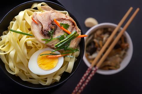 검은 접시에 고기와 달걀 조각이 있는 국수와 어두운 테이블에 젓가락이 달린 버섯 콩 채소의 전통적인 간식이 있는 딤섬 0명에