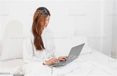 하얀 잠옷을 입은 여자가 주말 아침에 일어나 휴식을 취하고 노트북 휴대폰을 가지고 놀며 하얀 침실 안의 유리에서 빵을 먹고 차를 마신다 아침 휴가 개념입니다 갈색 머리에