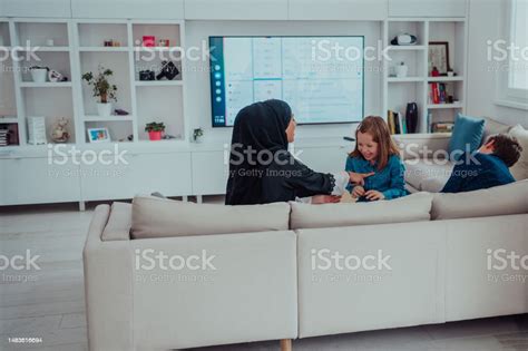 전통적인 유행 드레스를 입은 딸 여자와 함께 소파에 앉아 즐거운 시간을 보내는 행복한 무슬림 가족 2명에 대한 스톡 사진 및