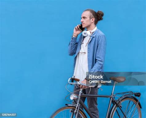 복고풍 스타일의 자전거를 들고 전화통화를 하는 파란색 벽 근처에 서있는 흰색 헤드폰을 입은 트렌디한 현대 청년 남자에 대한 스톡 사진 및 기타 이미지 Istock