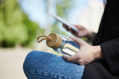 재사용 가능한 유리병을 들고 있는 젊은 여자 물병을 손에 들고 벤치에 앉아 있는 알아볼 수 없는 여성 리필에 대한 스톡 사진 및