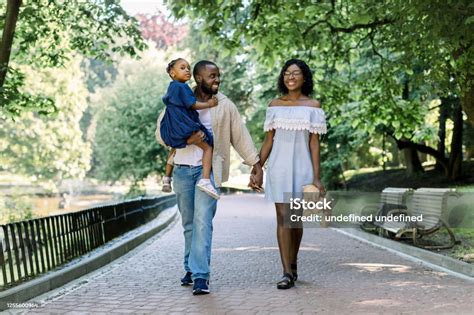공원에서 걷고있는 세 명의 사랑스러운 아프리카 가족의 전체 길이 초상화 잘 생긴 아버지는 팔에 자신의 딸을 잡고 그의 매력적인