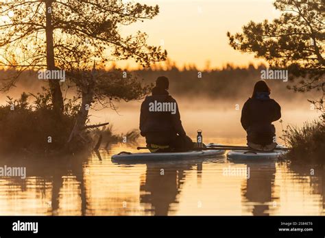 Two Paddleboarders Sit On A Calm Waterway With Mist Rising Golden