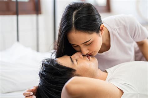 아시아인 아내는 침실에서 침대에 누워 있는 동안 남편에게 키스하고 있다 잠옷을 입은 아름다운 새 부부는 행복한 기분을 느끼고 이른 아침에 집에서 함께 여가 시간을 보내며