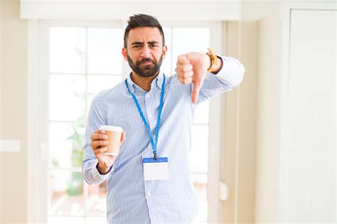 신분증을 쓰고 화가 난 얼굴로 커피 한 잔을 마시는 잘생긴 히스패닉 남자 엄지 손가락으로 싫어하는 것을 보여주는 부정적인 표지판