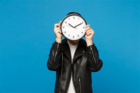 파란색 벽 배경 스튜디오 초상화에 고립 된 라운드 시계를 들고 검은 가죽 자 켓 흰색 T 셔츠에 잘생긴 세련 된 젊은 이루어지지 남자 사람들 라이프 스타일 개념입니다
