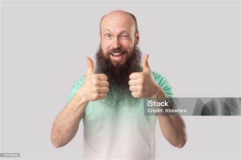 녹색 티셔츠에 긴 수염을 가진 기쁜 만족 중년 대머리 남자의 초상화 엄지 손가락 위로 윙크와 이빨 미소와 카메라를보고 40 49