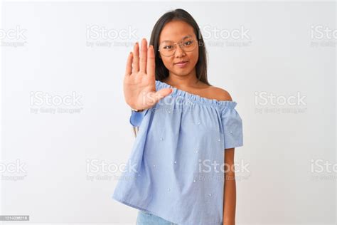 파란색 티셔츠와 안경을 쓰고 고립된 흰색 배경 위에 앉아 있는 젊은 중국 여성이 손바닥으로 노래를 멈추고 있다 얼굴에 부정적이고