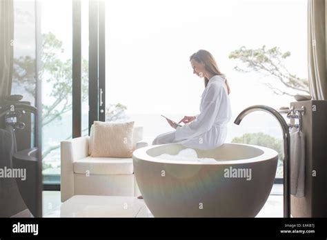 Woman Wearing White Bathrobe Sitting On Edge Of Modern Bathtub And