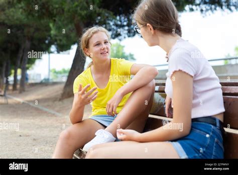 Two Girls Friends Sitting On Bench In Summer Park Chatting Happily