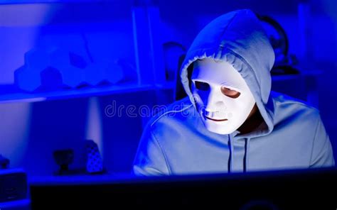 Male Hacker Wearing Mask And Hoodie Sitting In Dark Room Looking At