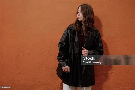 검은 가죽 재킷을 입은 젊은 여성이 왼쪽을 바라보며 주황색 벽 앞에 서 있는 모습을 보며 공간을 복사합니다 도시 스타일과 거리